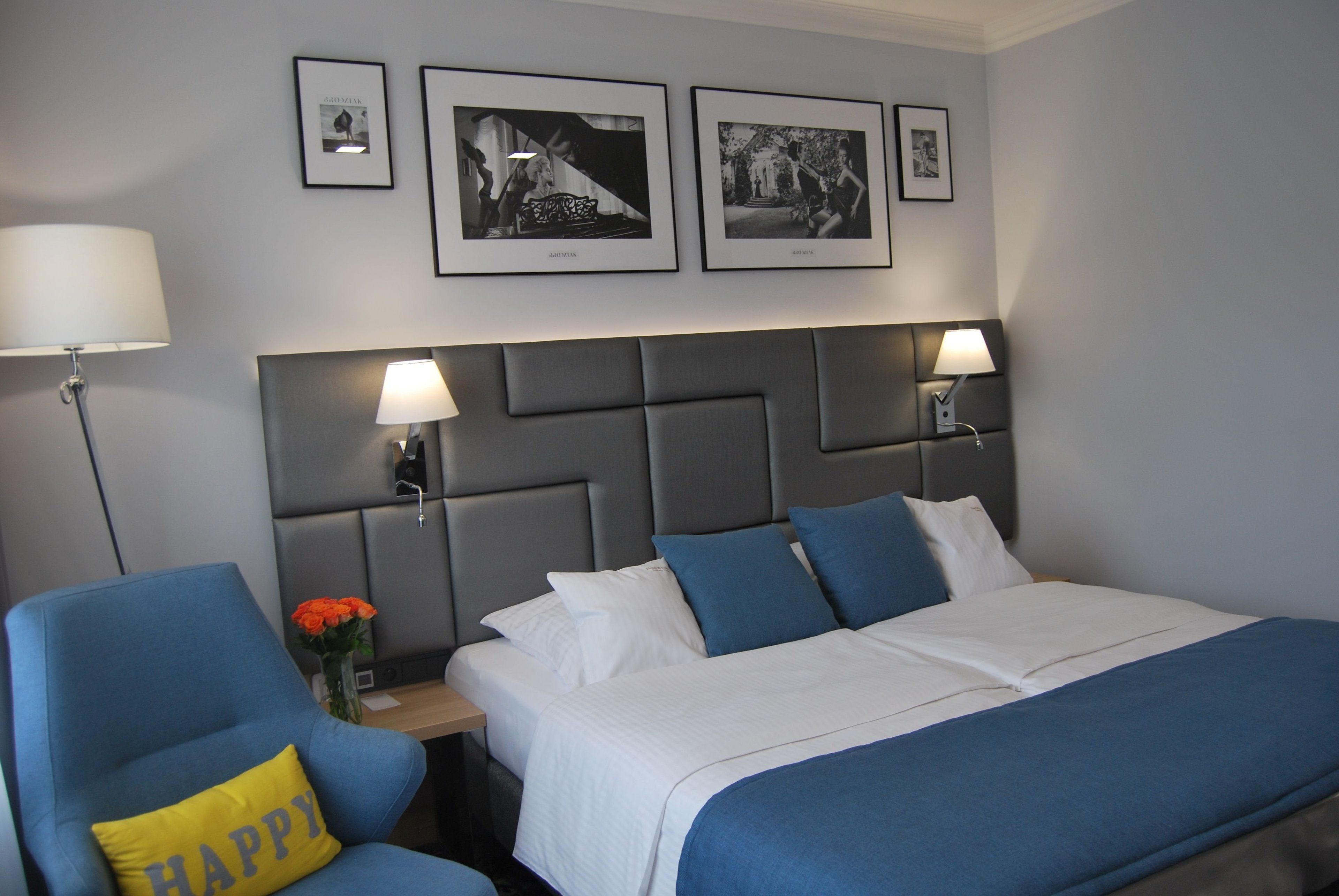 Quarto de hotel com uma cama, duas mesas de cabeceira, uma cadeira com um travesseiro que diz 'HAPPY' e três quadros na parede.