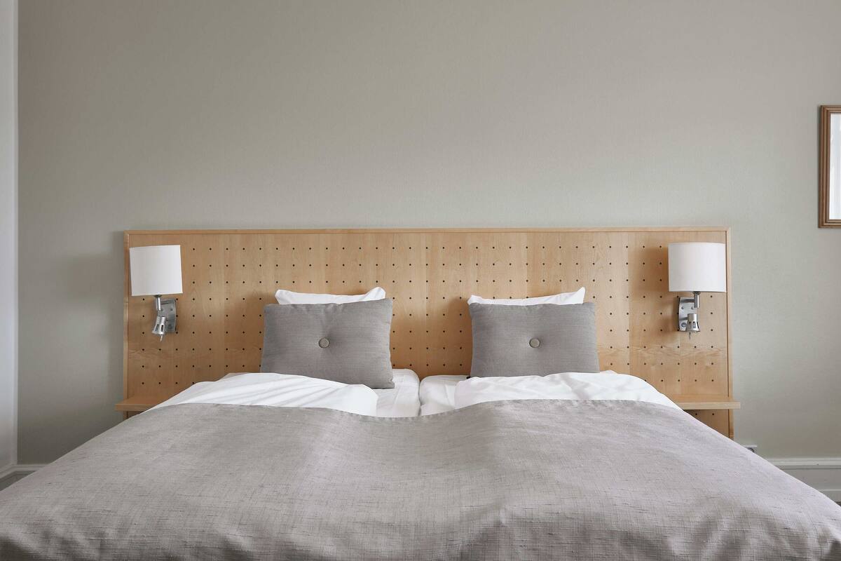 Cama bem arrumada com cabeceira de madeira, dois travesseiros e duas luminárias fixadas na parede.