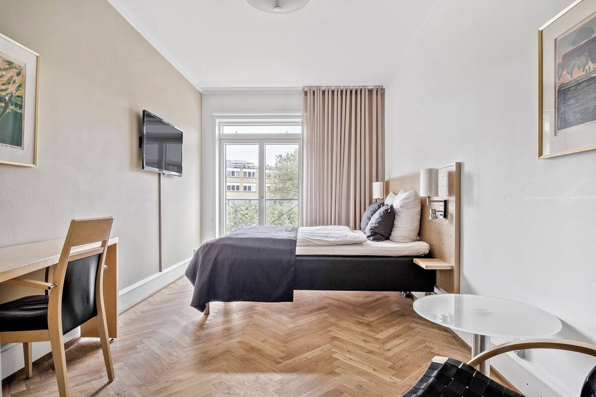 Quarto de hotel moderno com piso de madeira em padrão herringbone, cama com cabeceira escura, pequena mesa branca, escrivaninha com cadeira, TV fixada na parede e janela com cortinas.