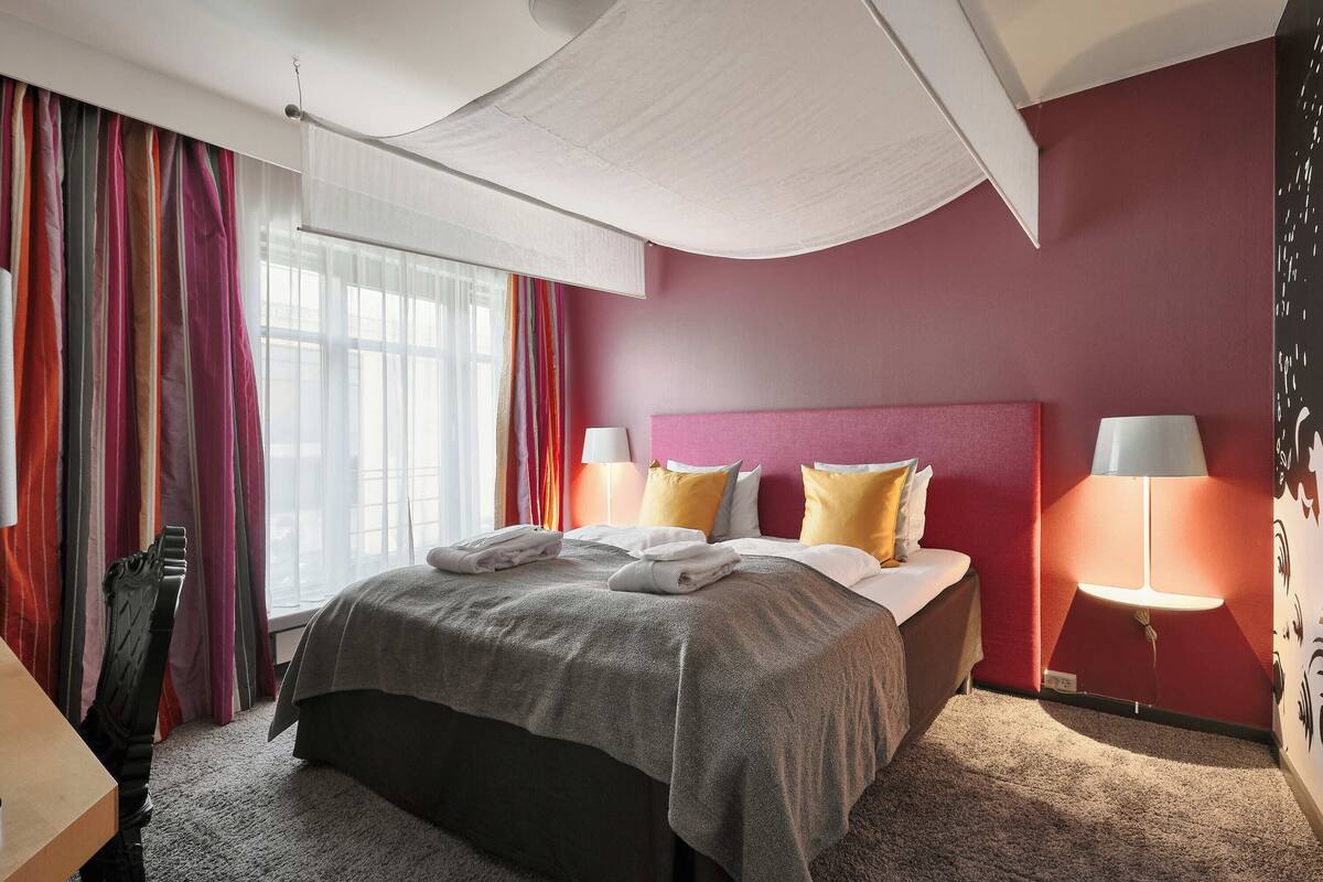 Quarto de hotel com cama grande, duas luminárias laterais e janela com cortinas.