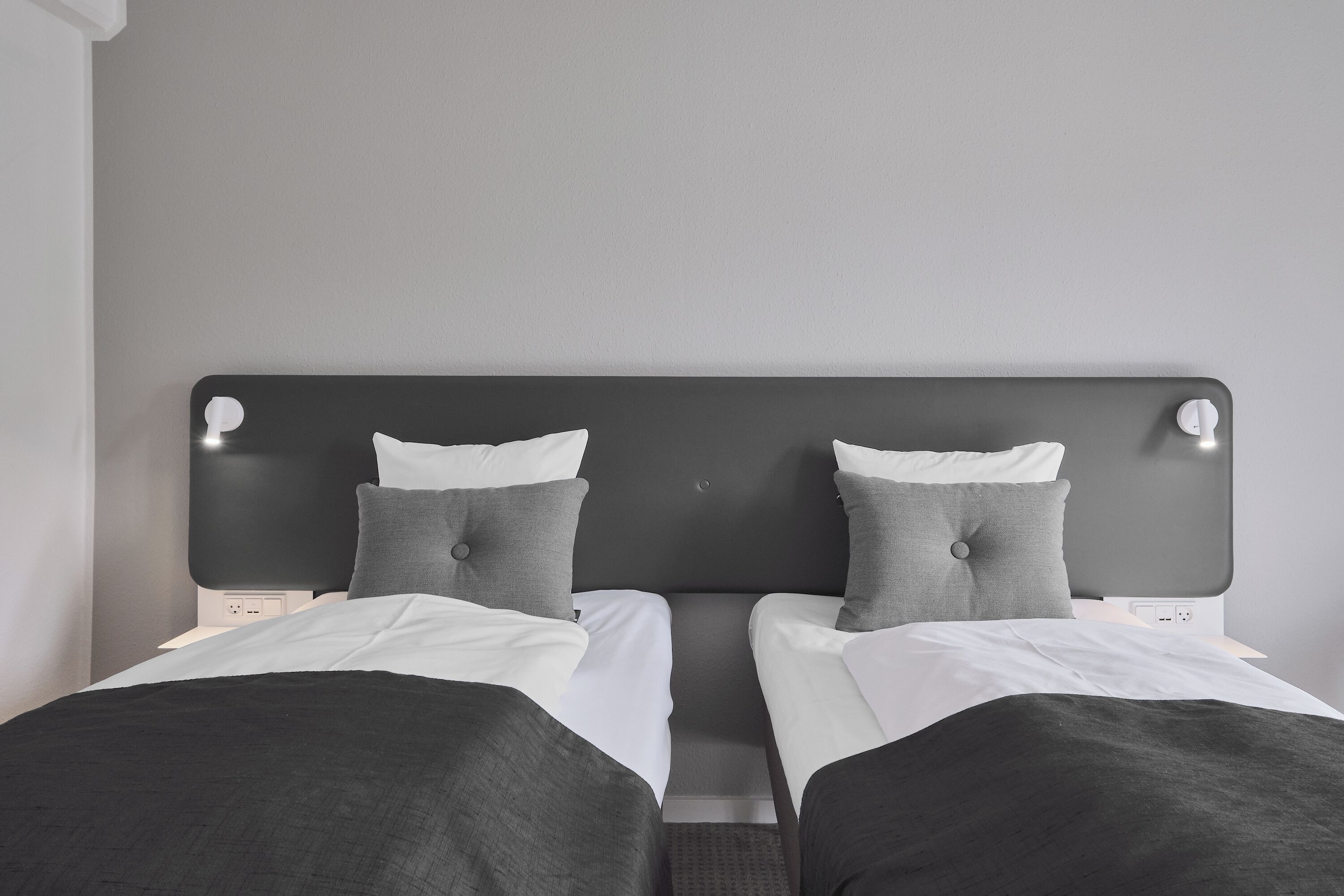 Cama de casal com cabeceira escura e duas almofadas, mesa de cabeceira com abajur e parede em tom neutro.