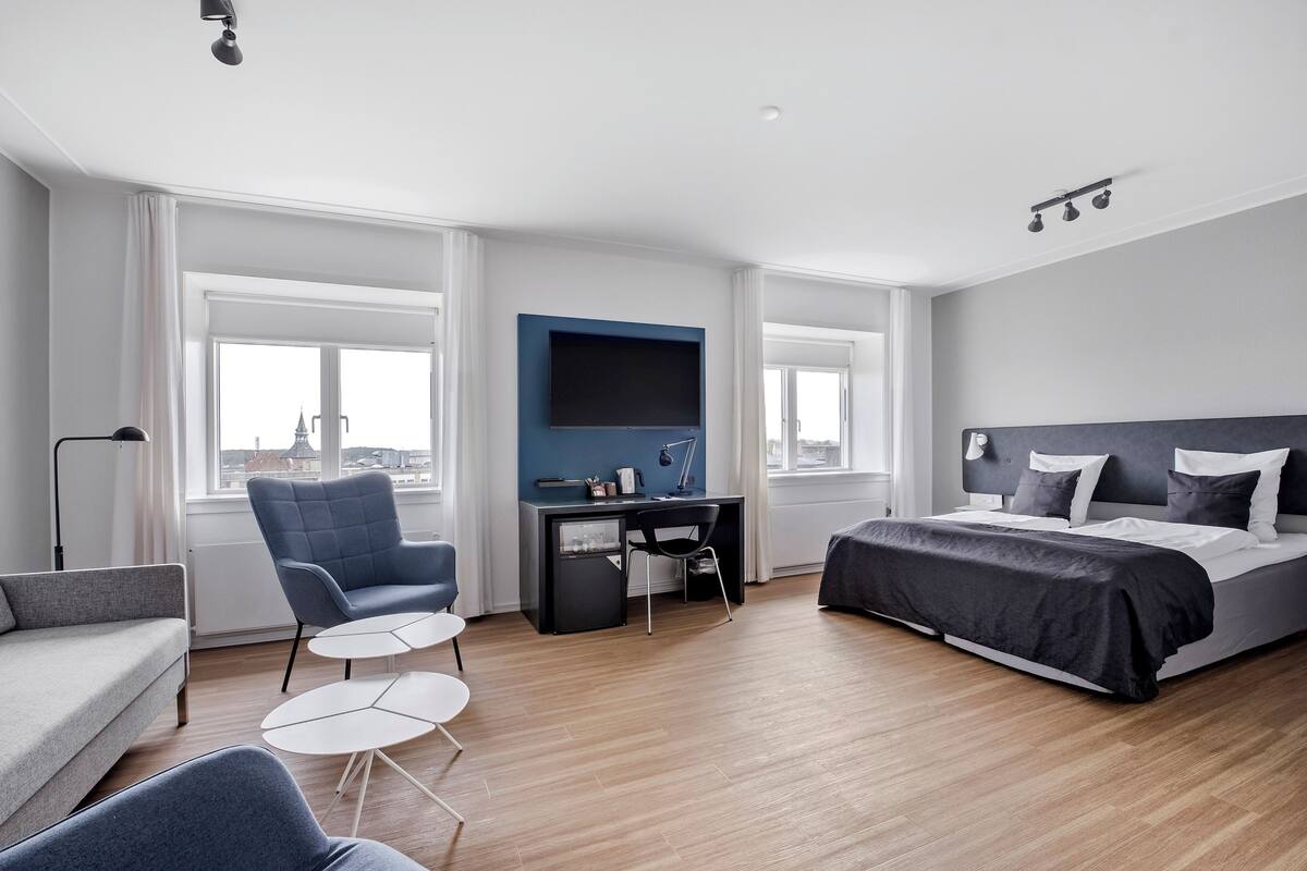 Quarto de hotel moderno com cama, escrivaninha, cadeira e duas mesinhas.