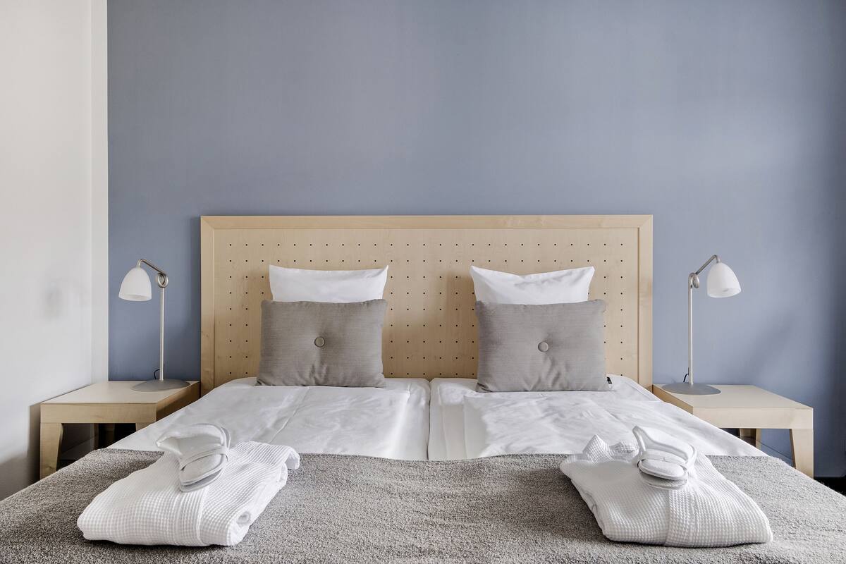Cama bem arrumada com duas almofadas, um cobertor cinza e duas toalhas dobradas em uma cabeceira de madeira.