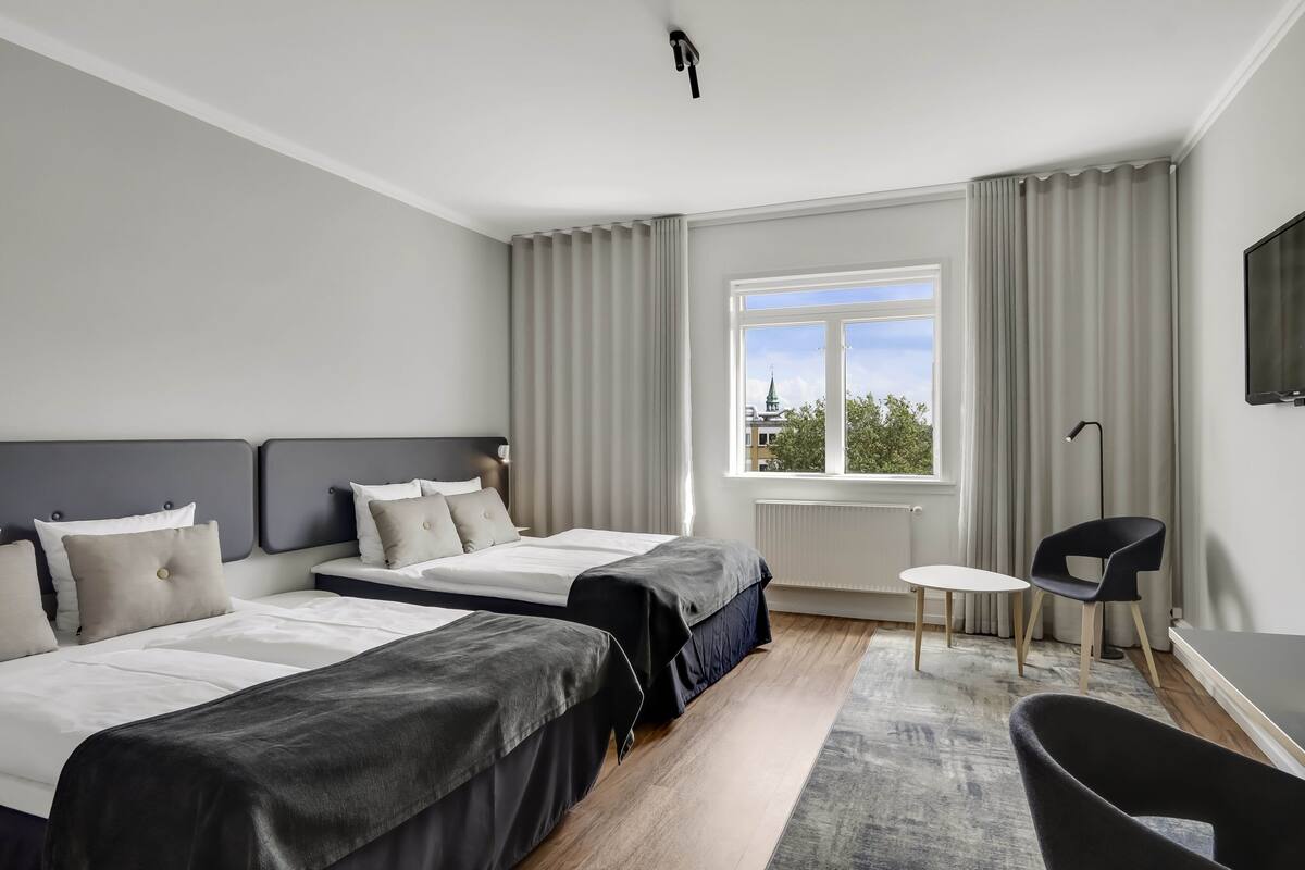 Quarto de hotel moderno com duas camas, uma mesa pequena, uma cadeira e uma janela grande com cortinas.