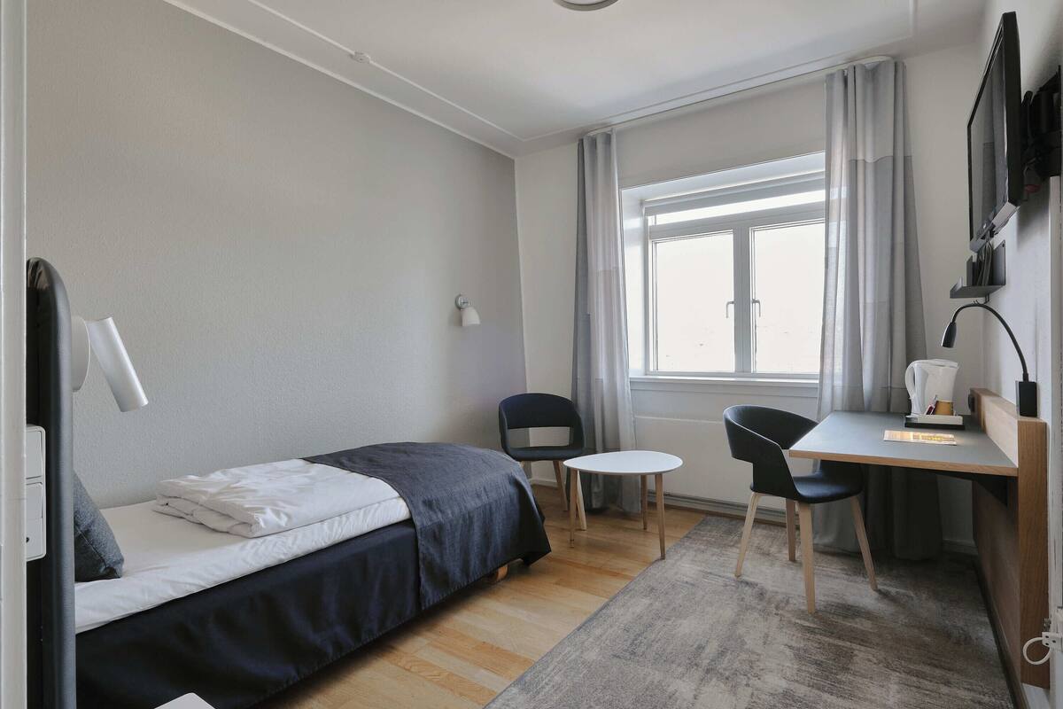 Quarto de hotel compacto com cama, escrivaninha, cadeira, mesa pequena, televisão e janela com cortinas.