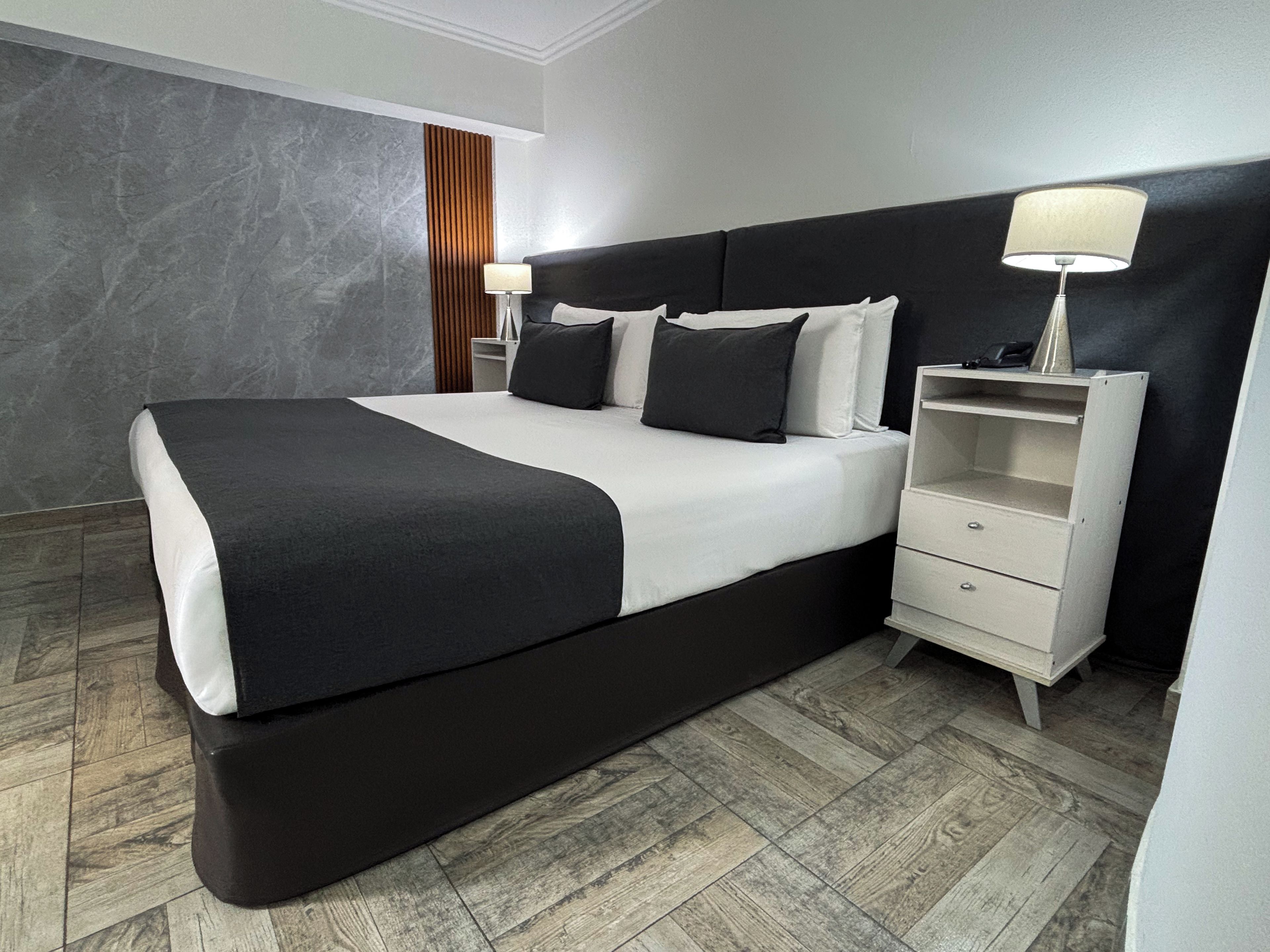 Quarto de hotel moderno com uma cama grande, uma mesa de cabeceira, um abajur e piso de madeira.