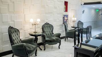 Um hall de hotel com design moderno, apresentando uma área de estar com cadeiras estofadas e uma mesa de vidro, piso de mármore e parede texturizada.