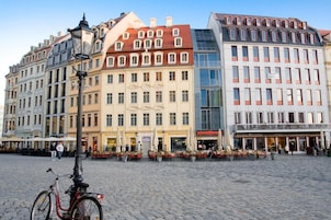 Uma praça de paralelepípedos com edifícios históricos, mesas ao ar livre e uma bicicleta.