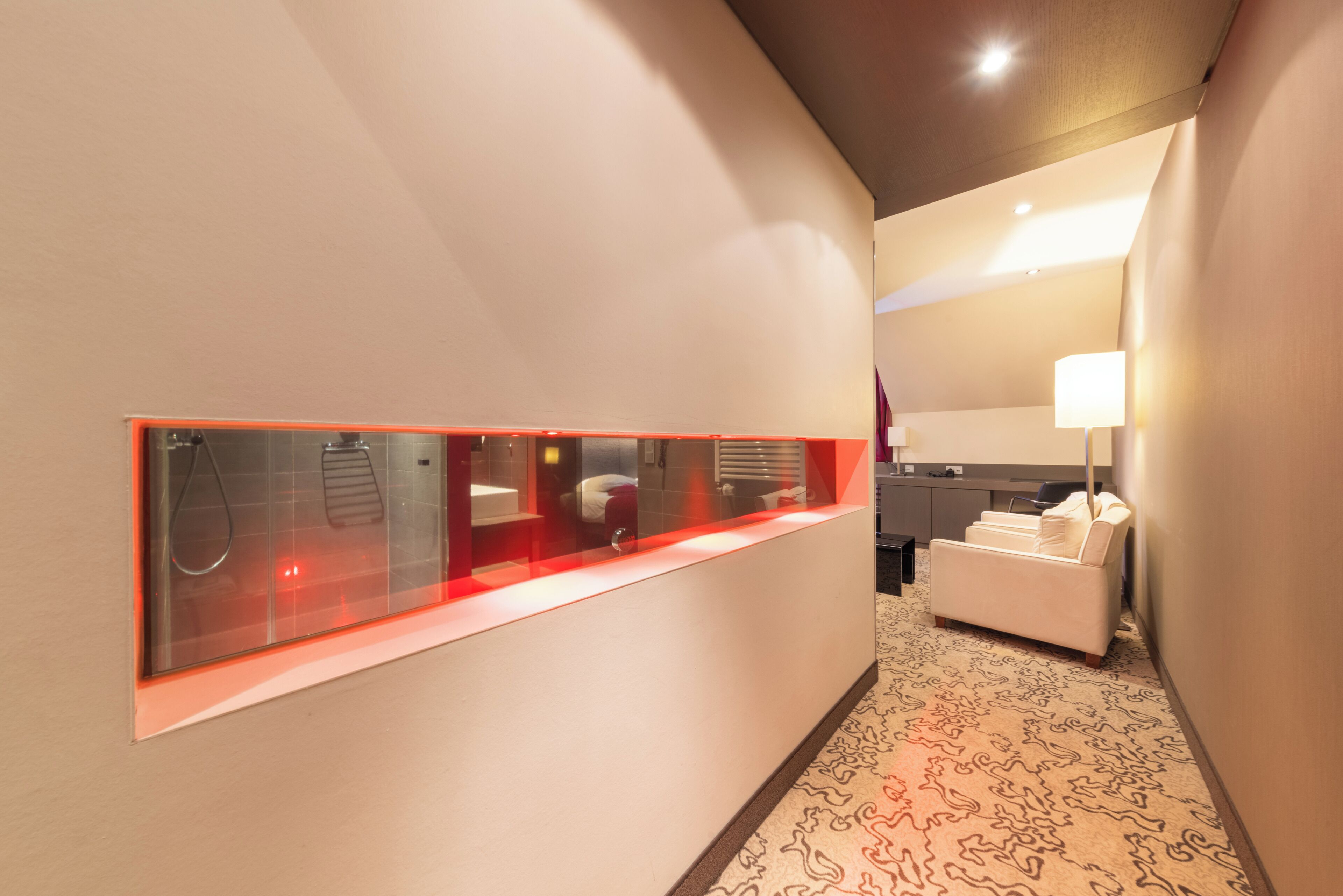 Um corredor de hotel com área de chuveiro em vidro, um sofá branco e uma luminária.