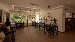 Uma ampla sala de jantar com piso de madeira, grandes janelas com vista para o exterior, e diversos conjuntos de mesas e cadeiras.