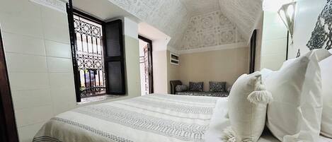Quarto com uma cama grande, colcha estampada, teto decorativo e janela com grade de metal.