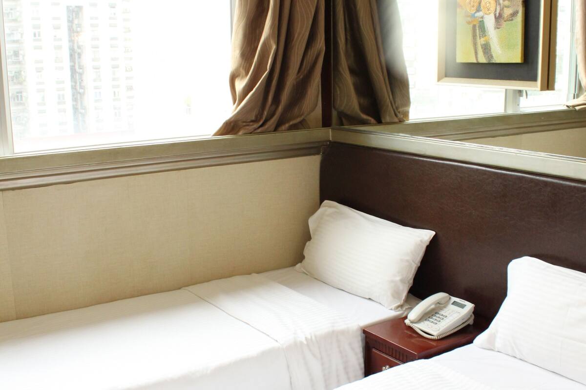 Quarto de hotel com uma cama de solteiro, mesa de cabeceira com telefone e janela com cortinas.