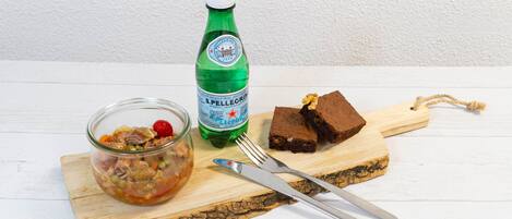 Uma tábua de corte de madeira com um pote de vidro de vegetais em conserva, uma garrafa de água S.Pellegrino e dois pedaços de bolo de chocolate.