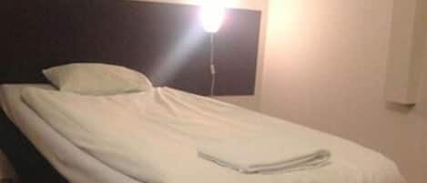 Uma cama bem arrumada com lençóis brancos e uma cabeceira escura em um quarto com parede branca lisa e piso de madeira.