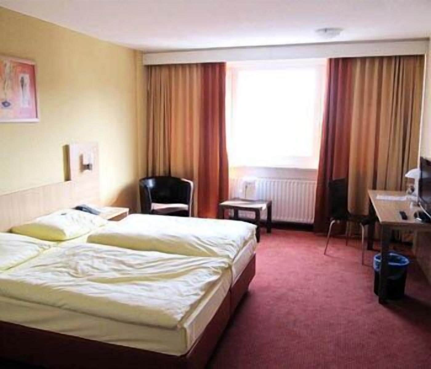 Quarto de hotel com cama, escrivaninha, cadeira e janela com cortinas.