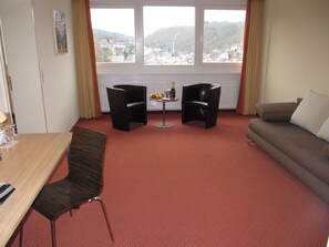 Quarto de hotel com uma mesa de jantar, duas cadeiras pretas, um sofá castanho e uma pequena mesa com uma garrafa e copos.