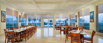 Uma ampla sala de jantar com várias mesas e cadeiras, grandes janelas com vista para palmeiras e uma piscina, e um design de teto moderno.