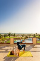 Uma pessoa fazendo uma pose de yoga em um tapete amarelo, com vista para uma praia e palmeiras ao fundo.