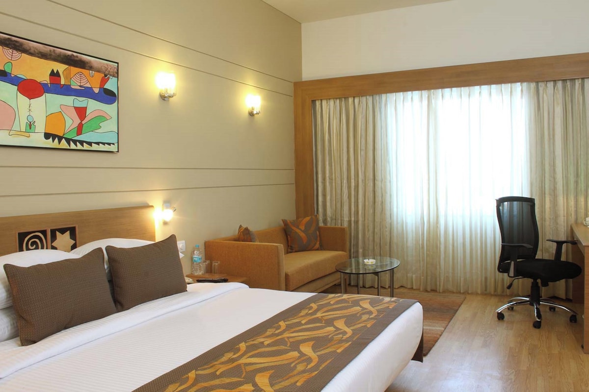 Quarto de hotel com cama, sofá, escrivaninha, cadeira e um quadro na parede.