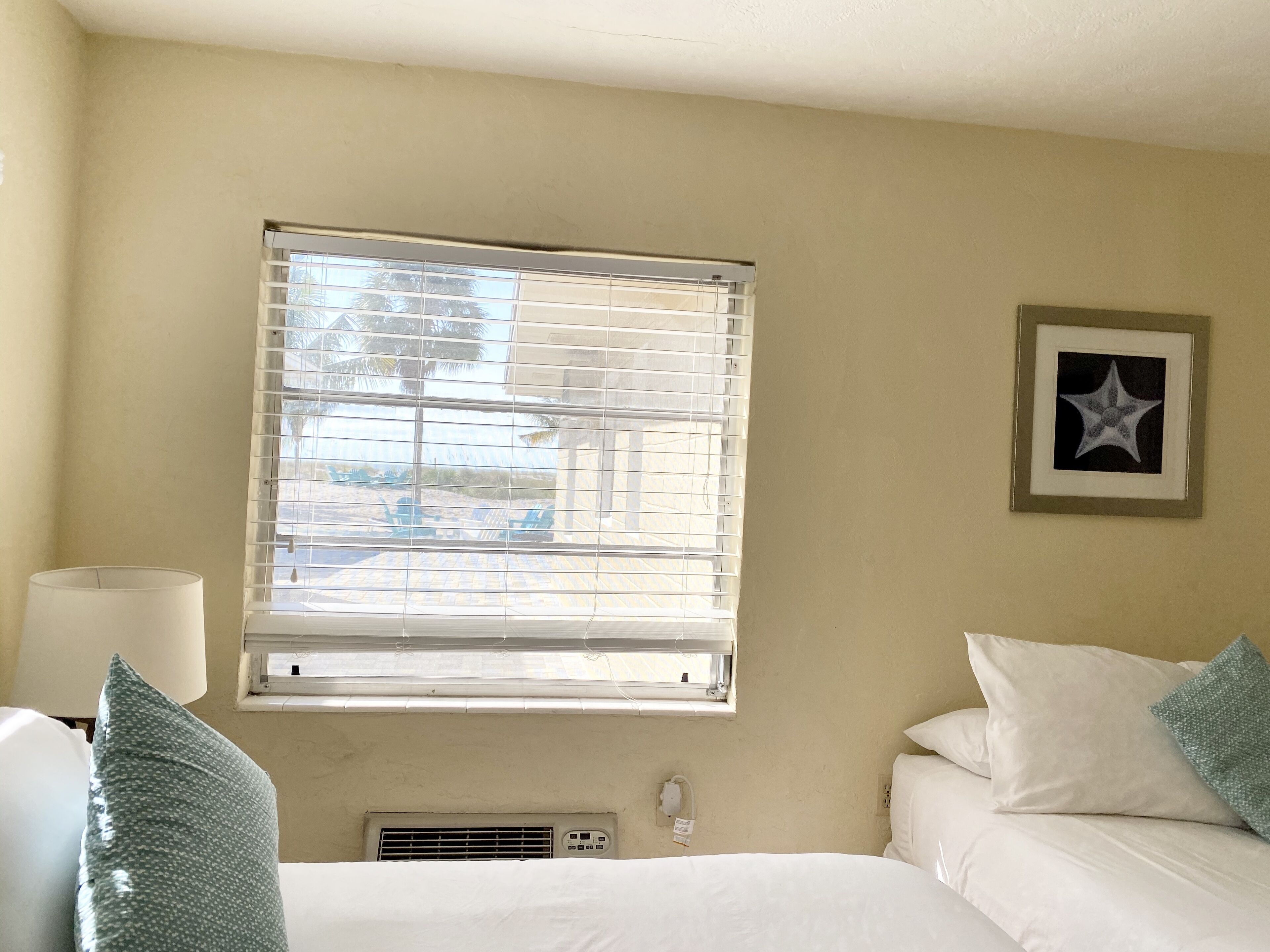 Quarto de hotel com cama, janela com persianas, ar condicionado fixado na parede e um quadro emoldurado.