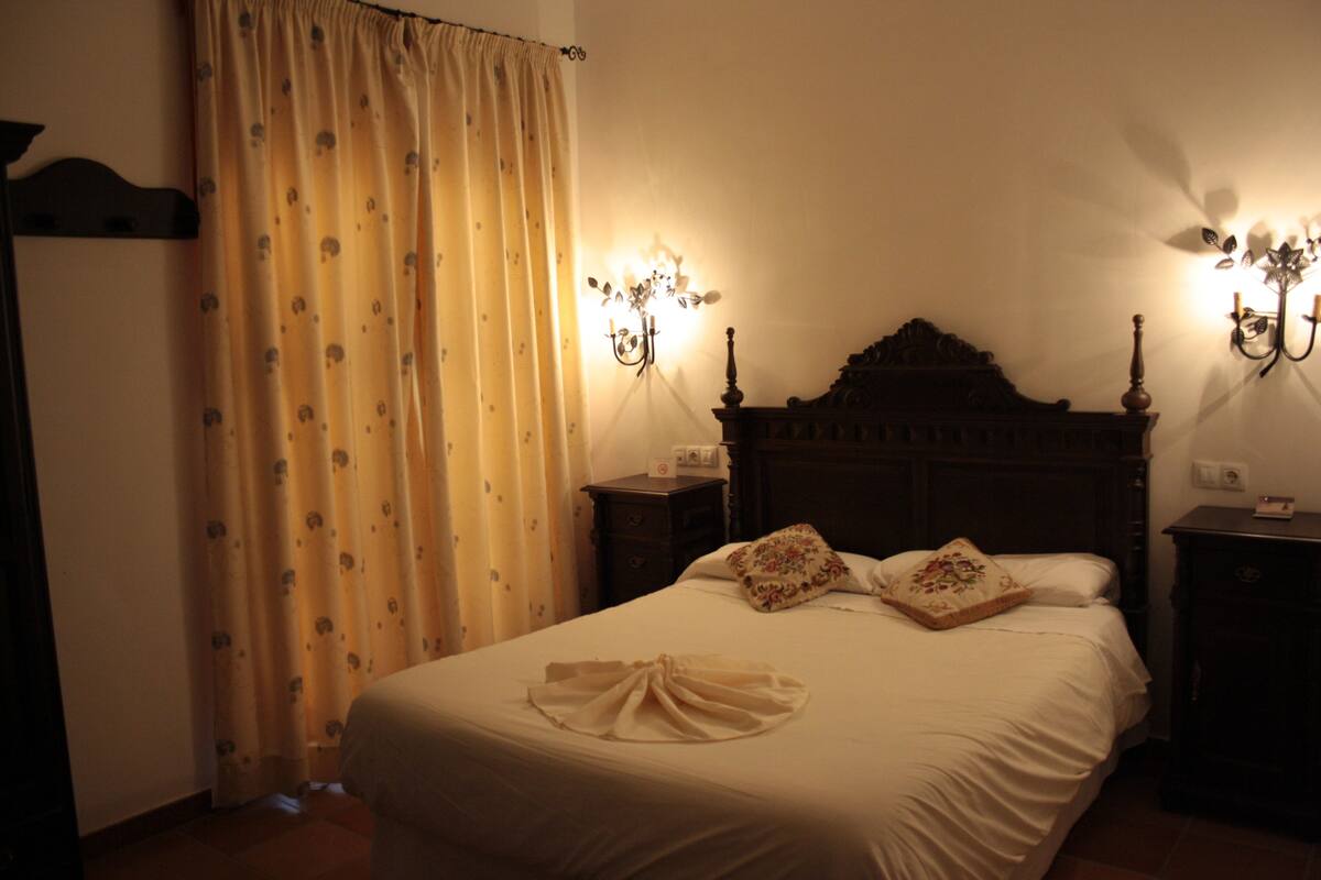 Quarto com cama de madeira, duas mesas de cabeceira, luminária na parede e cortina.