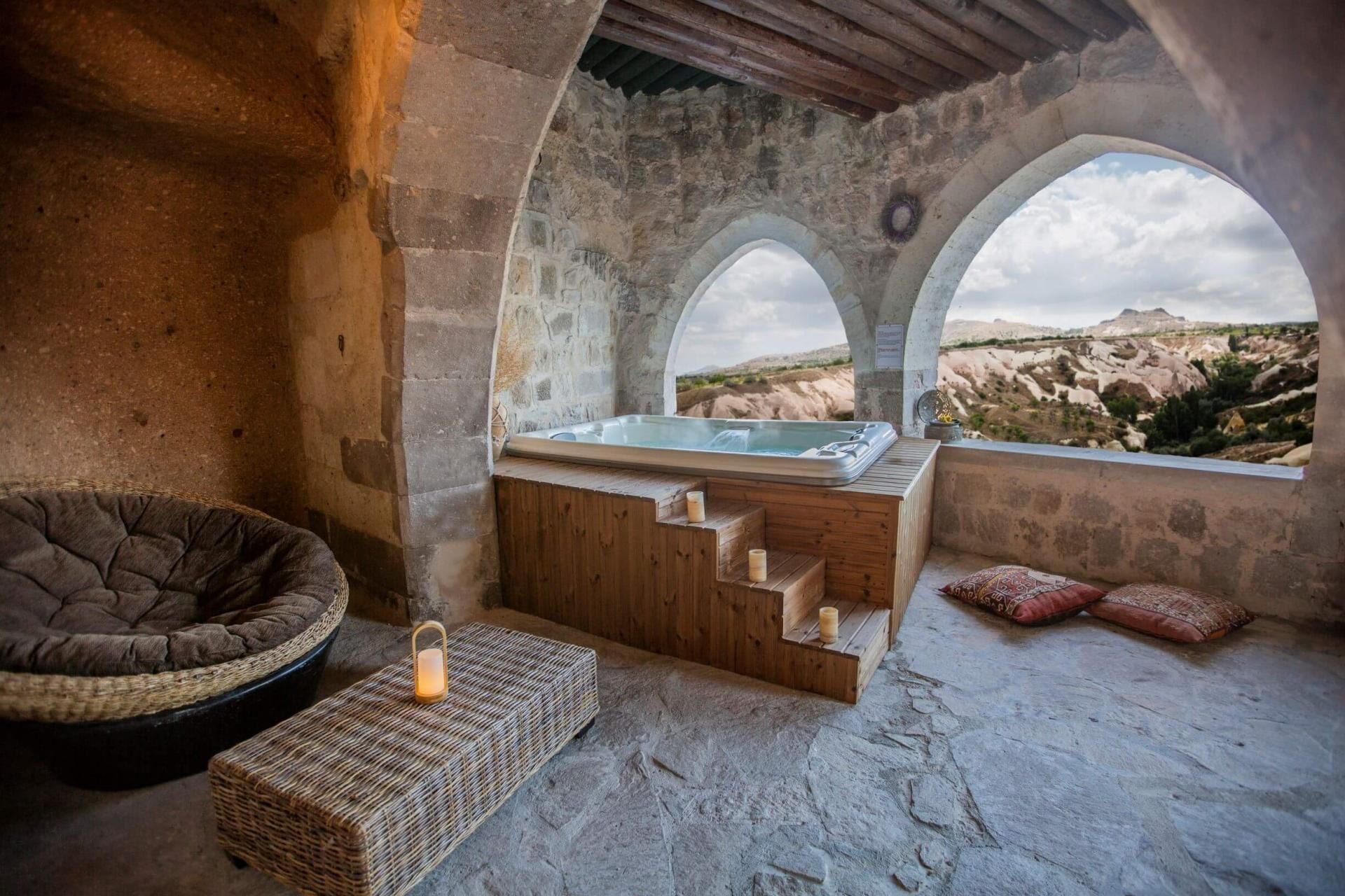Um quarto de paredes de pedra com um banco de madeira, uma pequena mesa com velas e vista para uma paisagem montanhosa através de janelas em arco.