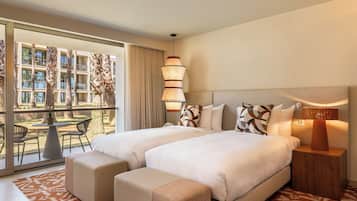 Quarto de hotel com duas camas, uma varanda com mesa e cadeiras, e vista para palmeiras.
