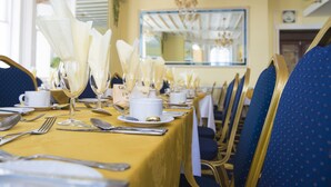 Uma sala de jantar com mesas preparadas para uma refeição formal, com pratos brancos, talheres e copos de vidro transparente.