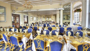 Um amplo salão de banquetes com mesas dispostas para refeições, com toalhas de mesa e cadeiras douradas e azuis.