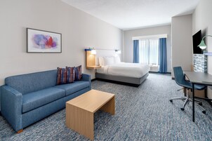 Quarto de hotel com um sofá azul, uma mesa de centro de madeira, uma cama com roupa de cama branca, uma escrivaninha com cadeira e um quadro na parede.