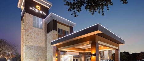 Entrada de hotel com design moderno, área de estacionamento coberta e céu claro.