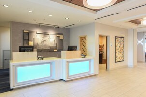 Uma área de recepção de hotel com design moderno, apresentando um balcão de atendimento com painéis iluminados, um espelho grande acima do balcão e uma câmera de segurança fixada na parede.