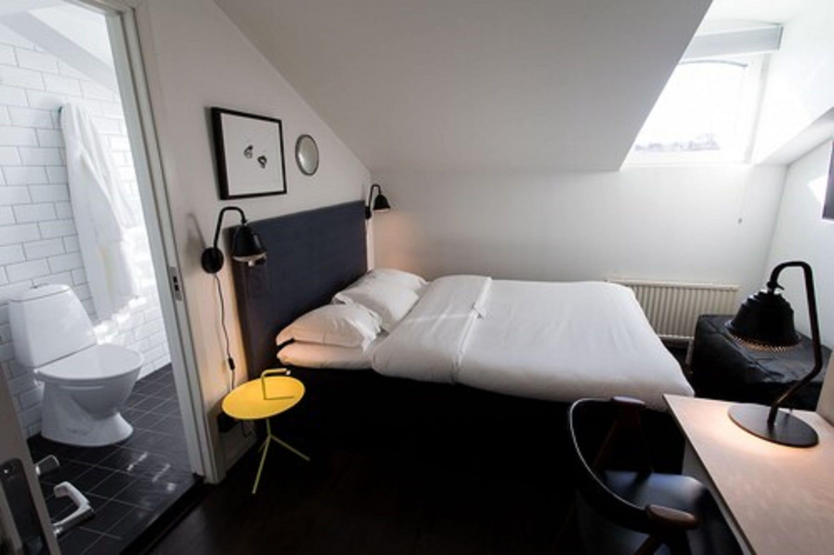 Quarto de hotel moderno com cama, mesa de cabeceira, cadeira e uma pequena mesa redonda.
