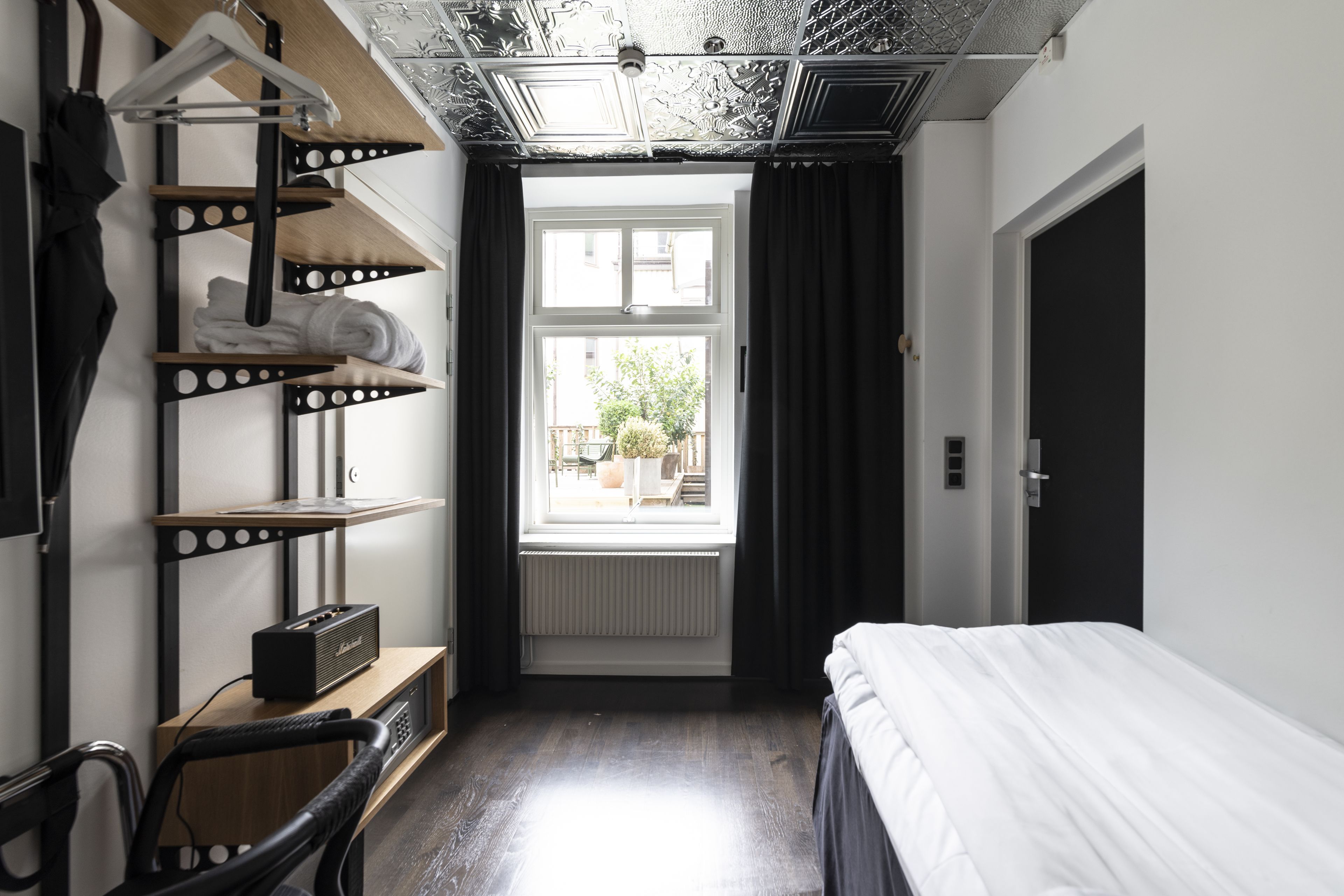 Quarto de hotel pequeno com uma cama, uma prateleira de madeira com toalhas, um radiador e uma janela com cortinas.