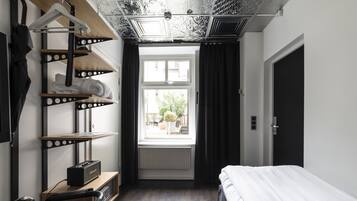 Quarto de hotel pequeno com uma cama, uma prateleira de madeira com toalhas, um radiador e uma janela com cortinas.