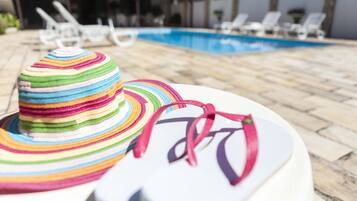 Um chapéu listrado e óculos de sol sobre uma superfície branca, perto de uma piscina com espreguiçadeiras ao fundo.