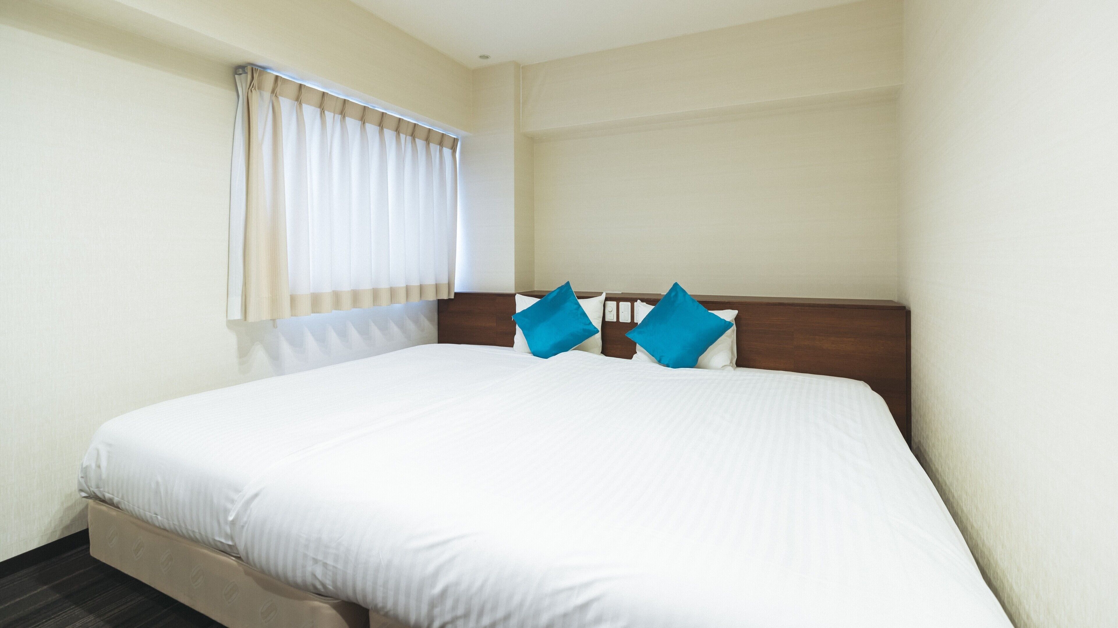 Quarto de hotel com uma cama grande, duas almofadas, uma janela com cortinas e uma cabeceira de madeira.