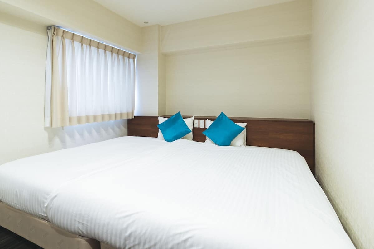 Quarto de hotel com uma cama grande, duas almofadas, uma janela com cortinas e uma cabeceira de madeira.