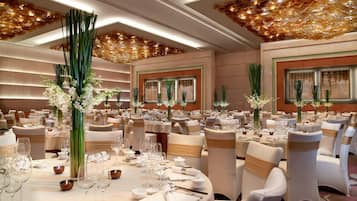 Uma sala de banquetes com mesas redondas, cadeiras brancas e centros de mesa florais.