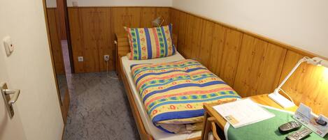 Quarto pequeno com uma cama de solteiro, painéis de madeira, colcha verde, luminária de cabeceira e dois controles remotos na cama.