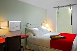 Quarto de hotel com cama, escrivaninha, cadeira, luminária, quadro emoldurado e banheiro com chuveiro.