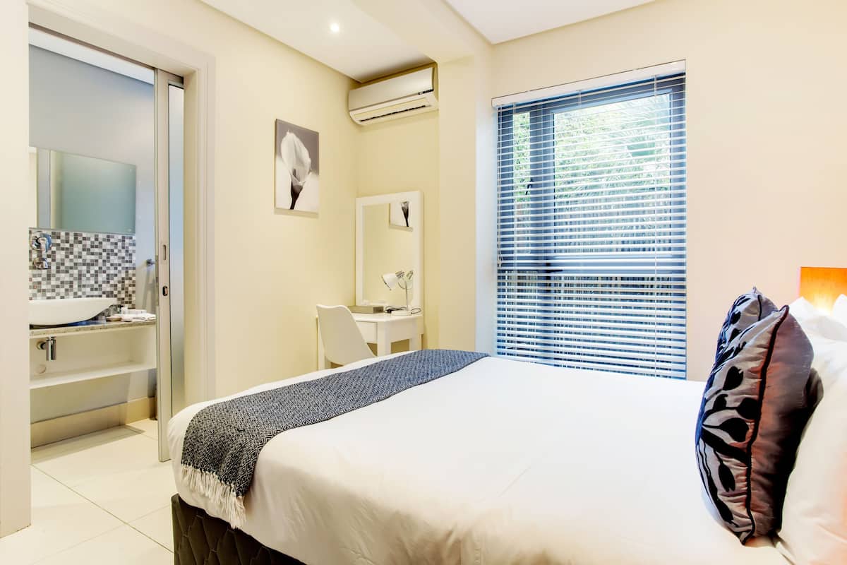 Quarto de hotel com cama, escrivaninha, cadeira, janela com persianas e banheiro com espelho e pia.
