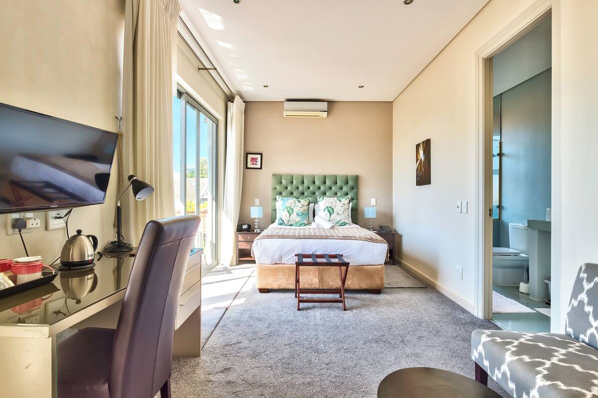Quarto de hotel com cama, escrivaninha com televisão, cadeira, mesinha e banheiro visível através de uma porta aberta.