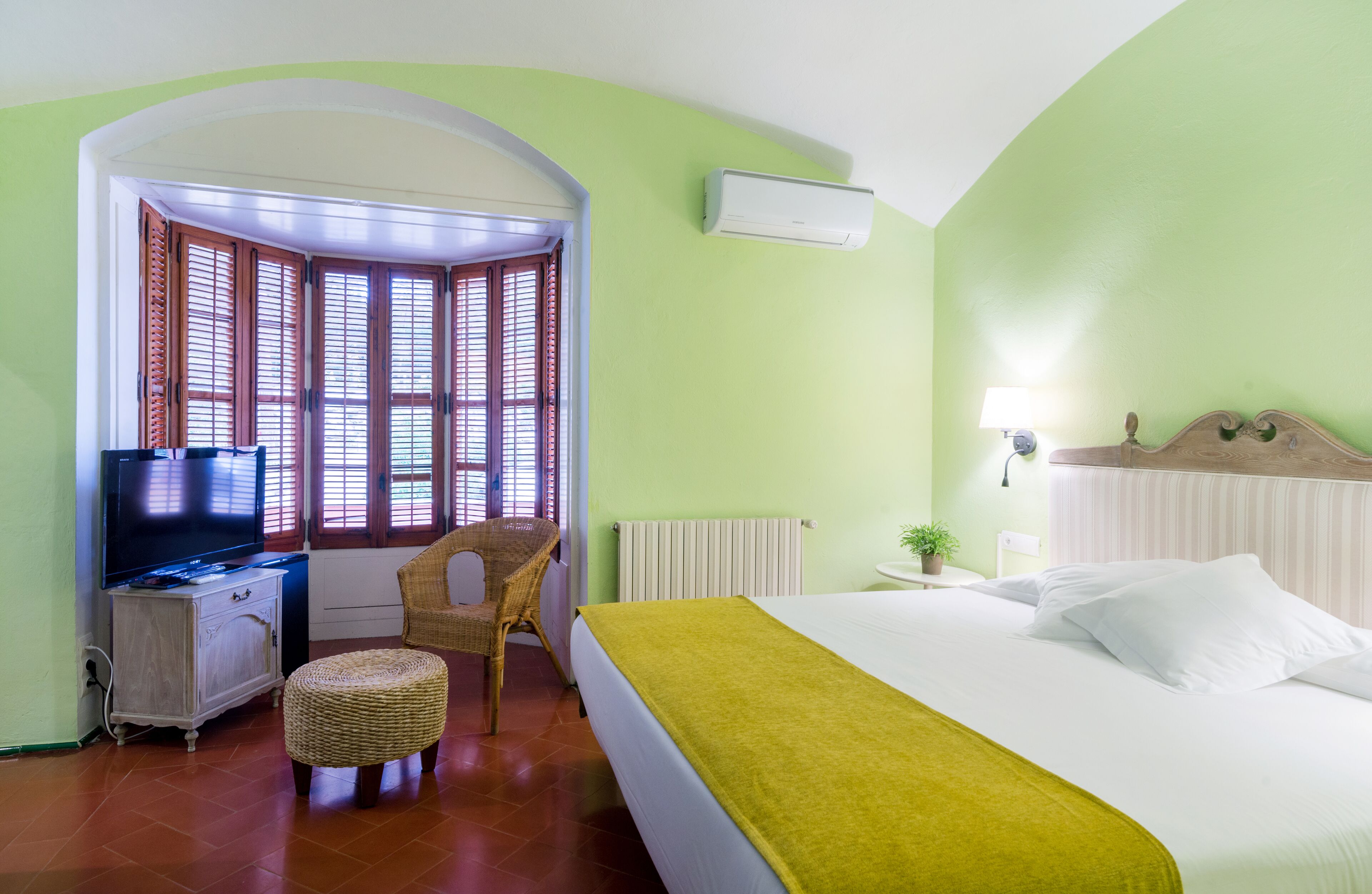 Quarto de hotel com uma cama, televisão, poltrona e janela com persianas.