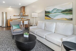 Uma sala de estar moderna com um sofá branco, uma mesa de centro preta e uma poltrona cinza. Uma área de cozinha é visível ao fundo, com uma geladeira, micro-ondas e armários de madeira.