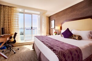 Quarto de hotel com uma cama grande, uma escrivaninha com cadeira, vista da cidade através de uma janela ampla e um tapete estampado.