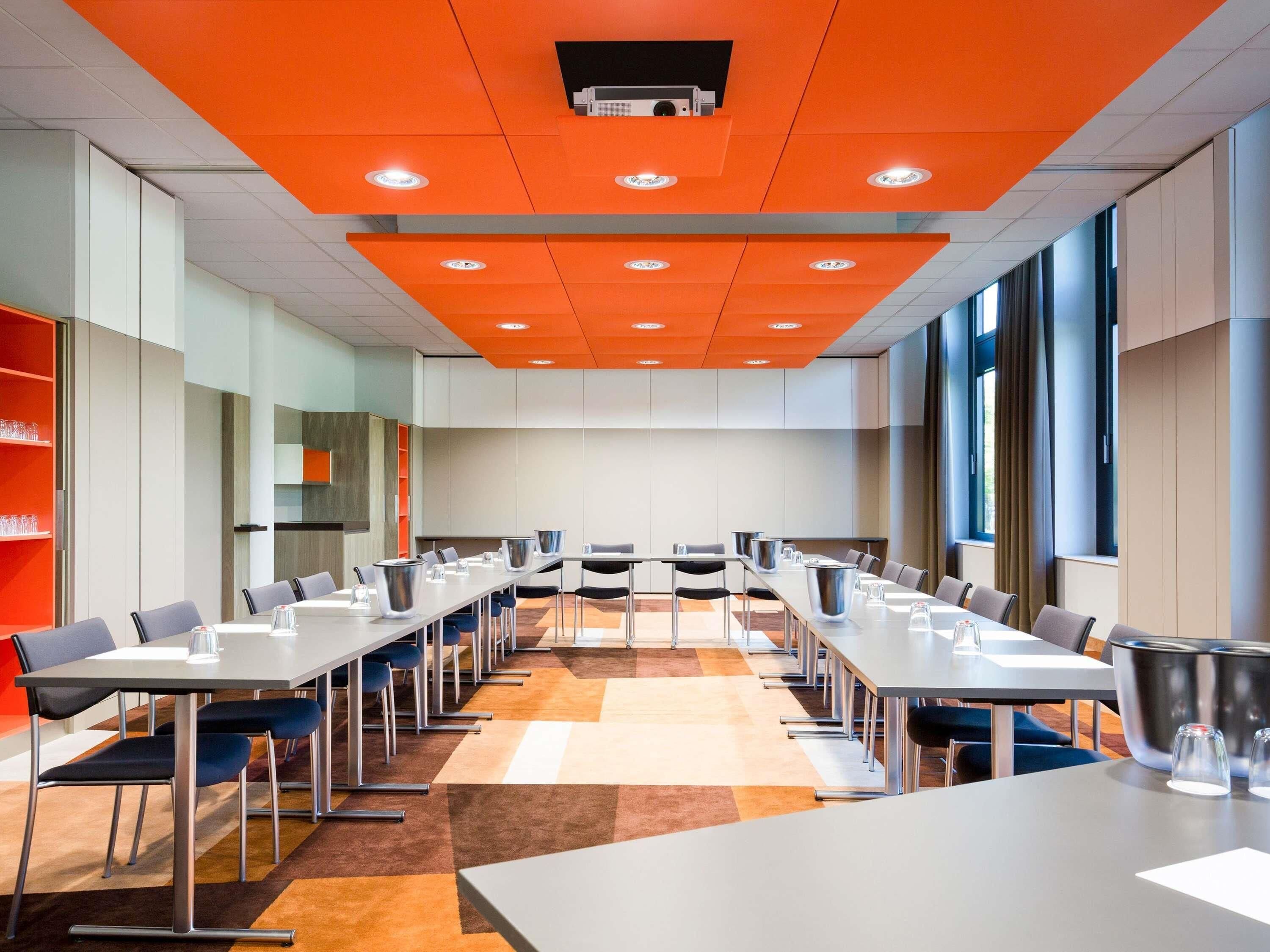 Uma sala de conferências com várias mesas e cadeiras, um projetor e grandes janelas.