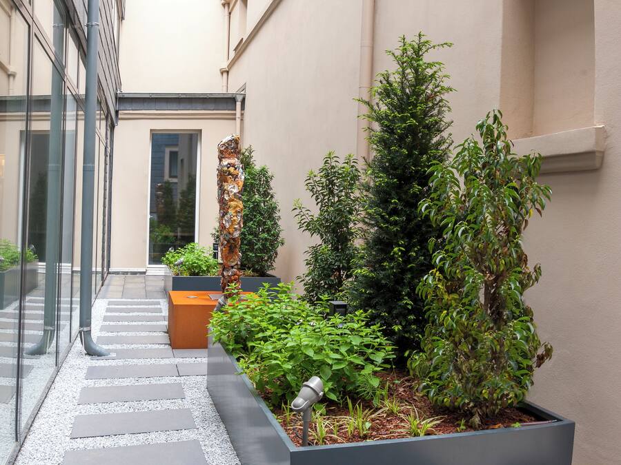 Um corredor exterior estreito com um painel de vidro de um lado e uma parede bege do outro. Há várias plantas em vasos e uma escultura decorativa alta no centro.