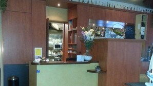 Uma área de recepção com balcão de madeira, um vaso de flores e uma placa indicando a carta de vinhos.