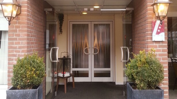 Entrada do Hotel Oranje Sittard, com uma placa sobre as portas, duas plantas em vasos de cada lado e uma pequena mesa com cadeiras em frente.